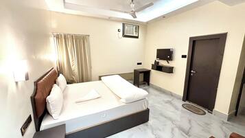 Quarto de hotel com cama, escrivaninha, ventoinha no teto, televisão e porta.