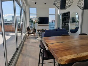 Um espaço de estar moderno com área de jantar, sofá, televisão e grandes janelas com vista para a cidade.