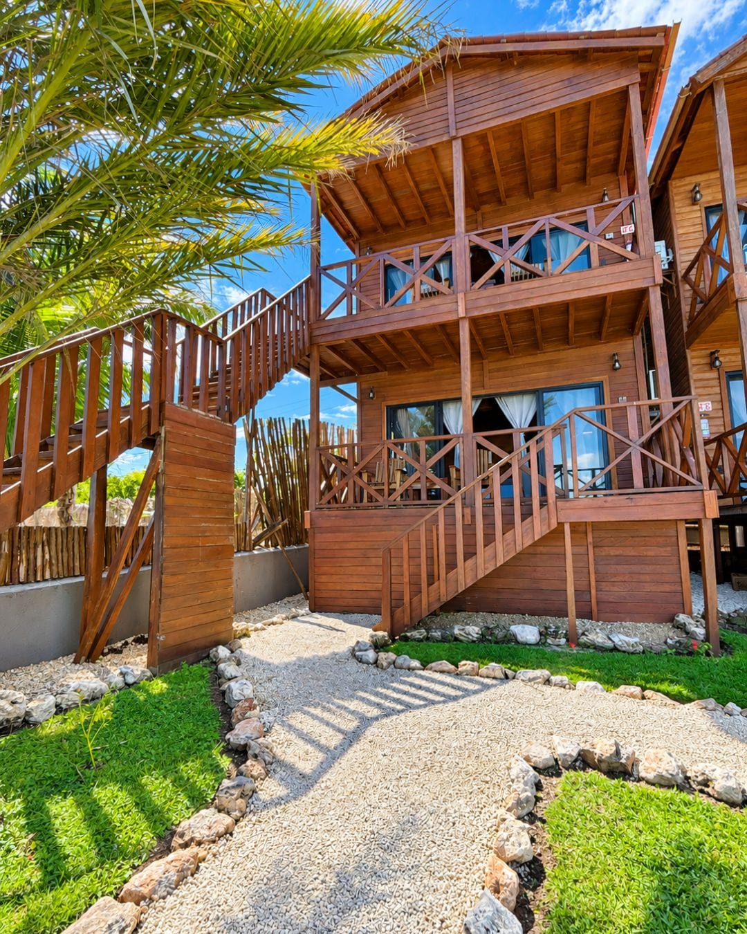 Cabana de madeira em vários níveis, com escada, cercada por um caminho de cascalho e vegetação.