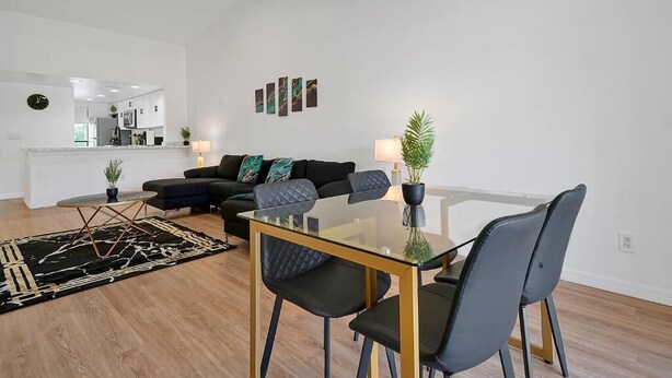 A modern living room with a glass dining table, black upholstered chairs, a black sofa with decorative pillows, and a black and gold patterned rug.