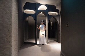 Uma pessoa de vestido branco em um corredor com portas arqueadas e iluminação embutida.