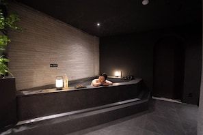 Uma área de spa com uma pessoa relaxando em uma banheira, uma luminária na parede e uma garrafa no balcão.