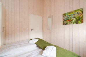 Uma cama de solteiro com roupa de cama branca e manta verde, uma porta branca e um espelho na parede.