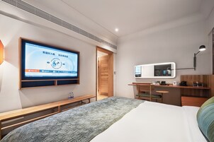 Quarto de hotel moderno com uma televisão de tela plana grande fixada na parede, um banco de madeira, uma cama com manta texturizada, uma escrivaninha com abajur e um espelho.