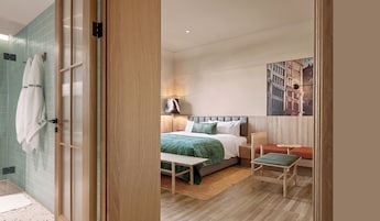 Quarto de hotel com cama, mesa de cabeceira, cadeira e um quadro na parede.