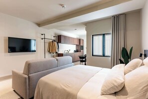 Quarto de hotel moderno com uma cama grande, área de estar, televisão de tela plana, cozinha compacta e janela com cortinas.