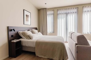 Quarto de hotel com uma cama, mesa de cabeceira, cadeira e janela com cortinas.