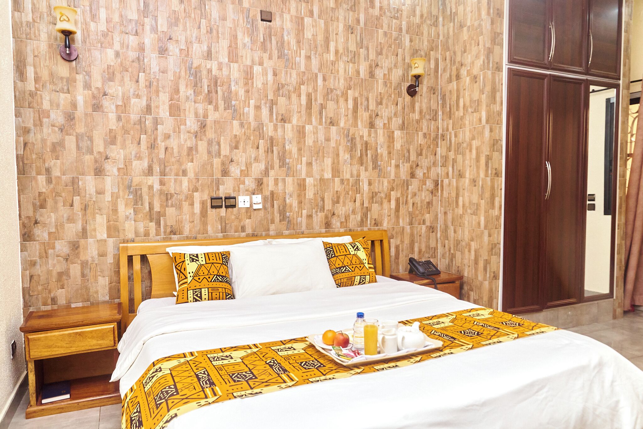 Quarto de hotel com parede com padrão de madeira, cama com colcha branca e amarela estampada, mesa de cabeceira com telefone e bandeja com itens de café da manhã.