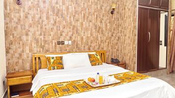Quarto de hotel com parede com padrão de madeira, cama com colcha branca e amarela estampada, mesa de cabeceira com telefone e bandeja com itens de café da manhã.