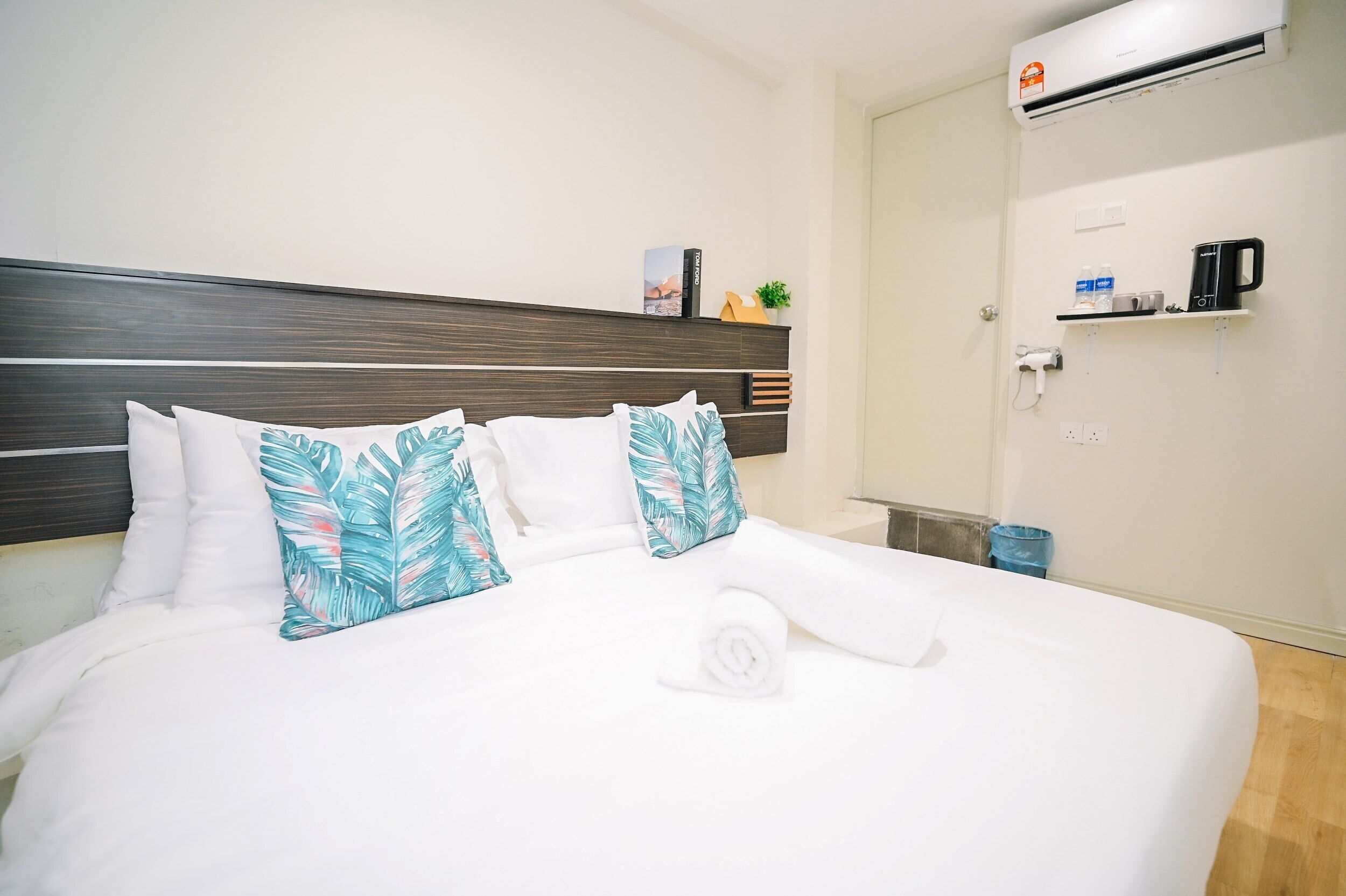 Quarto de hotel com cama, almofadas, toalha, ar condicionado, prateleira com artigos de higiene e uma porta.