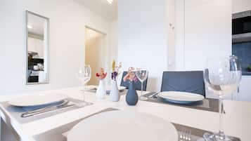 Uma área de refeições moderna com uma mesa branca posta para dois, incluindo pratos, talheres e copos. Há três vasos com flores secas sobre a mesa.
