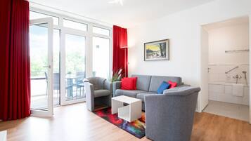 Uma sala de estar moderna com um sofá cinza, uma mesa de centro branca e um tapete colorido. Há uma área de jantar com uma mesa e cadeiras, e uma porta de vidro deslizante que dá para um espaço ao ar livre.