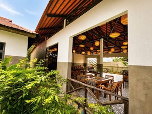 Uma área de refeições ao ar livre com mesas e cadeiras, um espaço de estar coberto com luminárias pendentes e a entrada de um edifício com um letreiro.