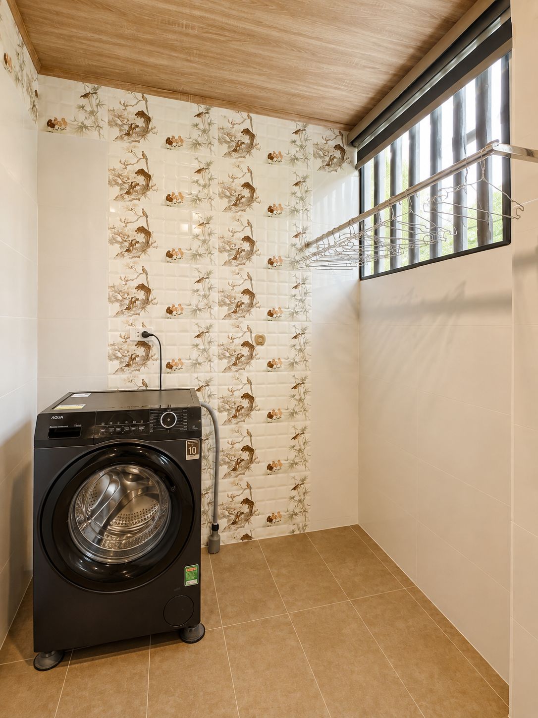 Uma lavanderia com máquina de lavar de abertura frontal, paredes revestidas com azulejos e um padrão inspirado na natureza, uma janela e um varal de metal, além de piso em azulejo.