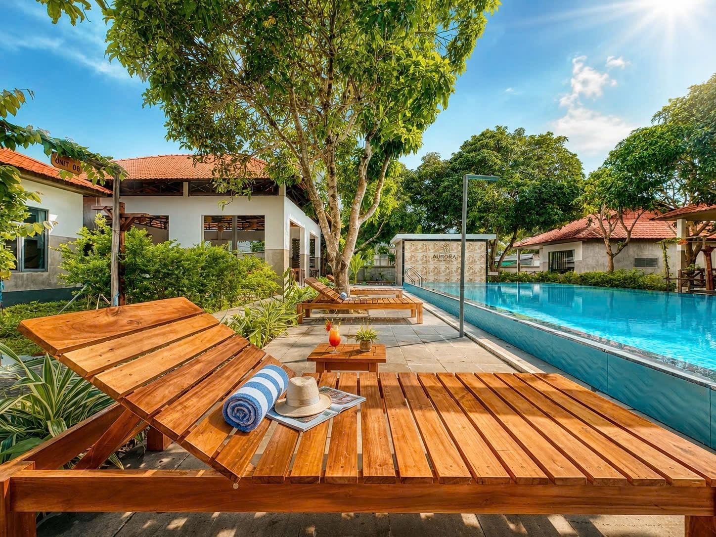 Uma área à beira da piscina com espreguiçadeiras de madeira, uma piscina e um edifício com a placa 'AURORA'.