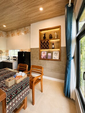 Espaço de refeições com mesa posta para dois, cozinha ao fundo e prateleira com objetos decorativos.