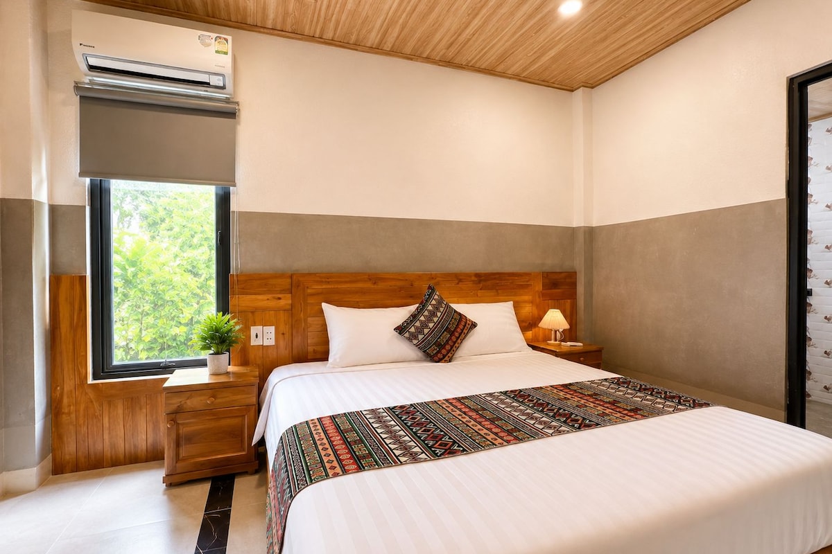 Quarto com cabeceira de madeira, cama com roupa de cama branca, manta estampada, mesa de cabeceira com abajur e janela com vista para o verde.