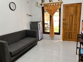 Um quarto com sofá cinzento, porta de madeira, janela com cortinas, relógio de parede e frigorífico.