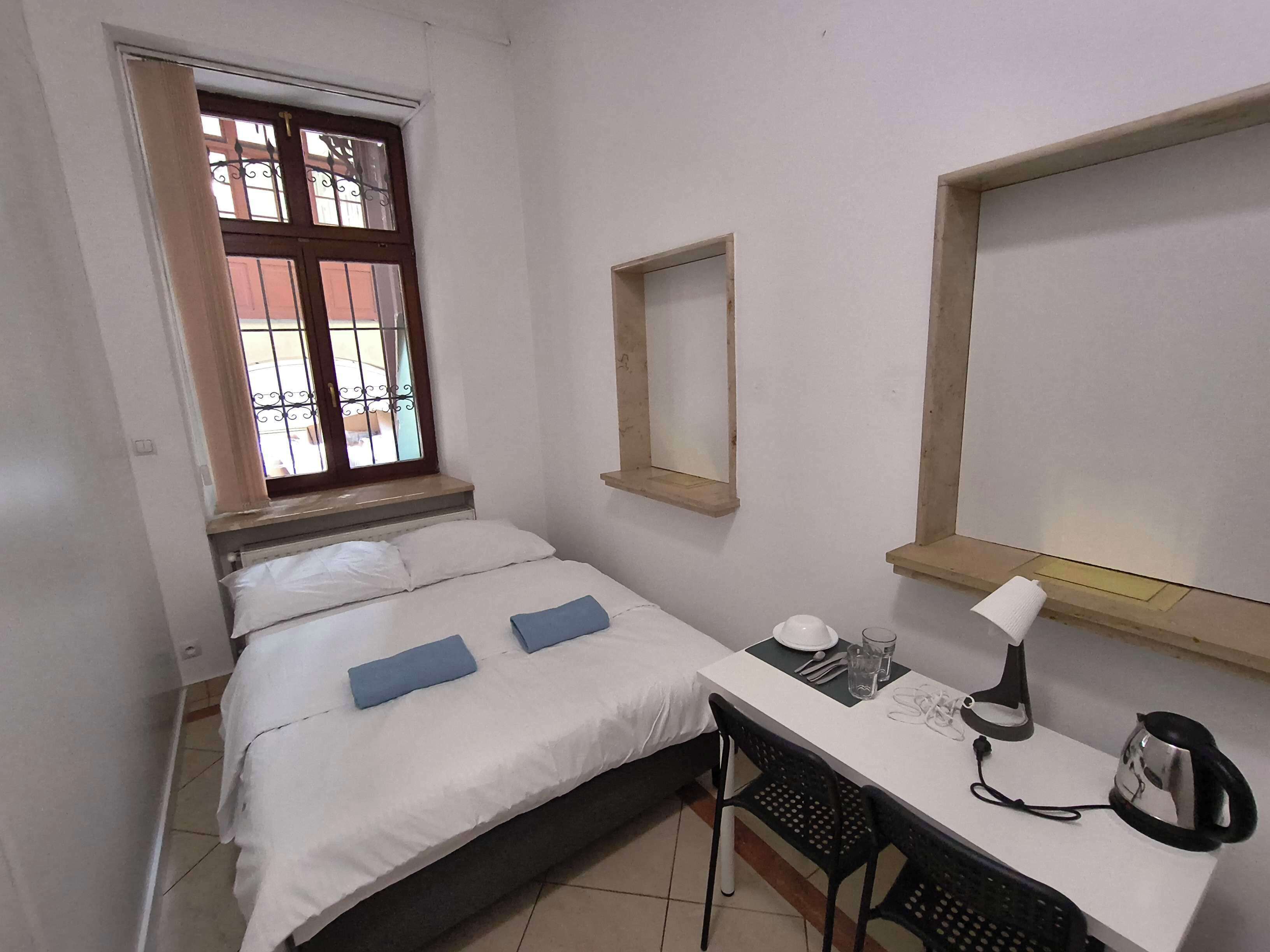 Um quarto pequeno e limpo, com uma cama, uma secretária com uma chaleira e uma janela com grades.