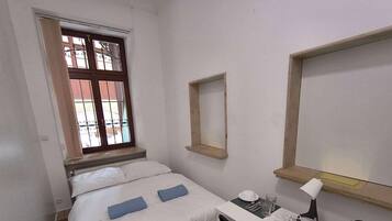 Um quarto pequeno e limpo, com uma cama, uma secretária com uma chaleira e uma janela com grades.