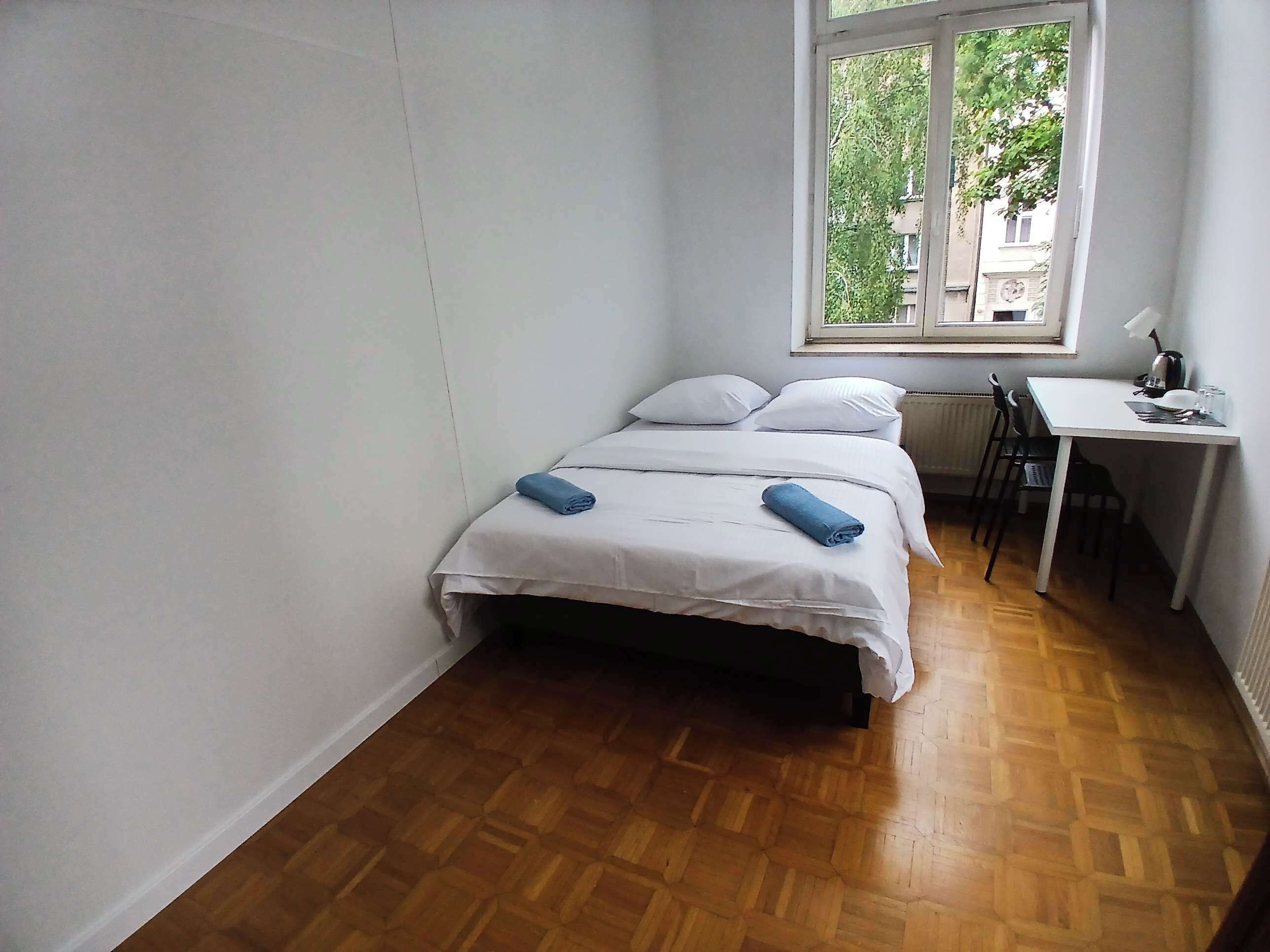 Quarto pequeno e limpo com cama, escrivaninha e cadeira. Uma janela permite a entrada de luz natural.