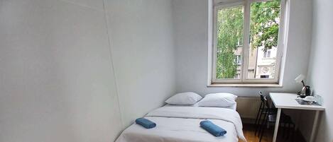 Quarto pequeno e limpo com cama, escrivaninha e cadeira. Uma janela permite a entrada de luz natural.