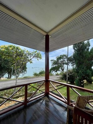 Uma área externa coberta com um deck de madeira, corrimão e vista para o mar e árvores.