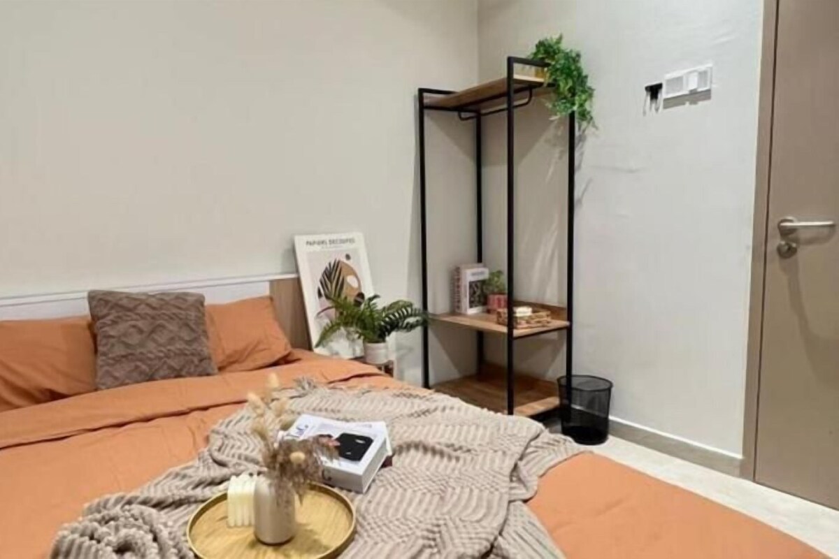 Quarto com cama, almofadas, mesa de cabeceira, prateleira com plantas e uma porta.