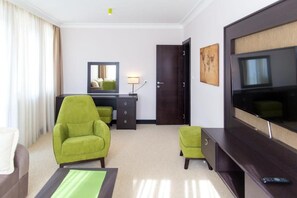 Quarto de hotel com uma poltrona verde, um banquinho pequeno, uma escrivaninha com abajur, uma televisão e uma porta.