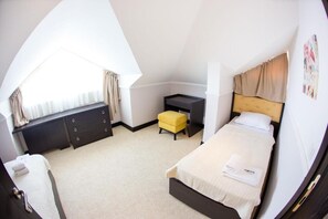 Quarto de hotel com cama, escrivaninha, cadeira, um banquinho pequeno e janela com cortinas.