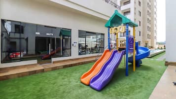 Parquinho infantil com escorregadores e estrutura de escalada em frente a um edifício com grandes janelas e varanda.