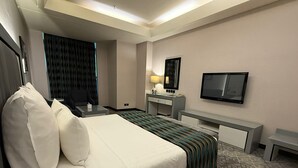 Quarto de hotel com cama, mesa de cabeceira, abajur, televisão e secretária com cadeira.