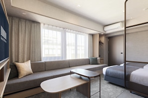Quarto de hotel moderno com sofá, mesa pequena, cama e janela grande com cortinas.