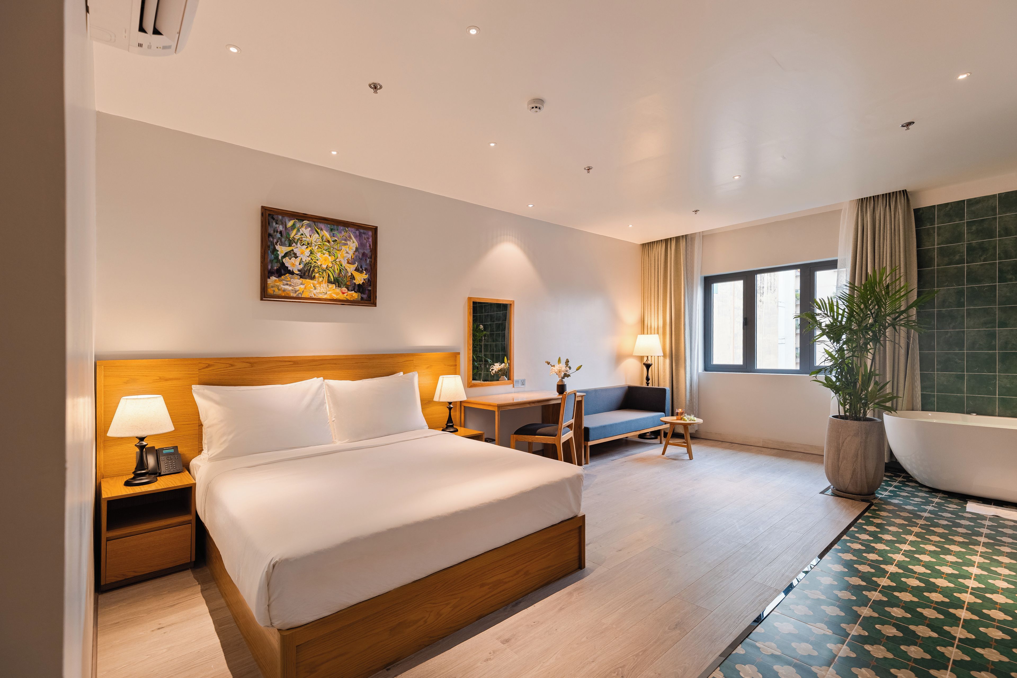 Quarto de hotel com cama grande, escrivaninha, poltrona, sofá, banheiro com banheira e uma janela ampla.