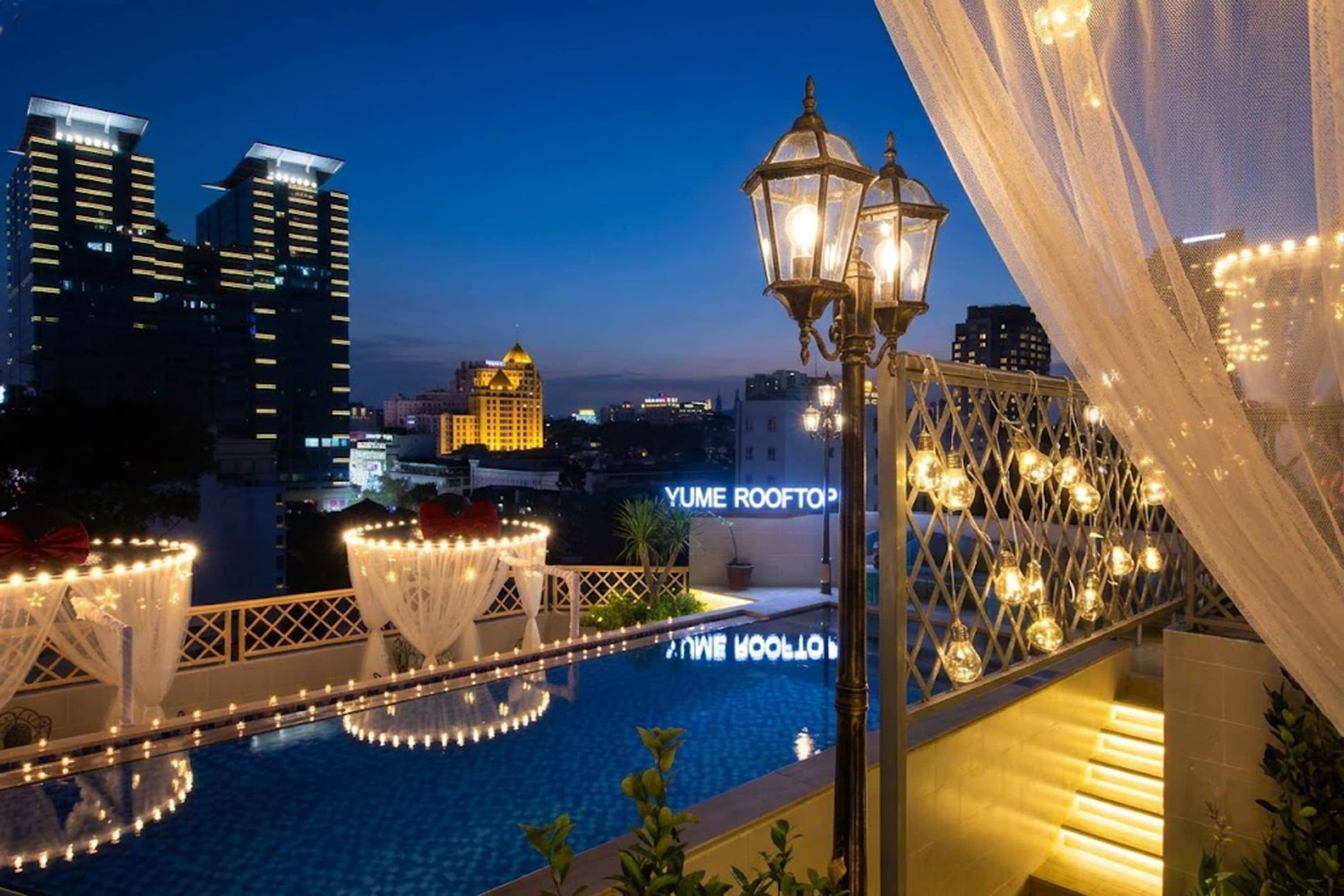 Piscina no terraço iluminada por luzes, com vista para o horizonte da cidade, e um letreiro com a inscrição 'VUME ROOFTOP'.