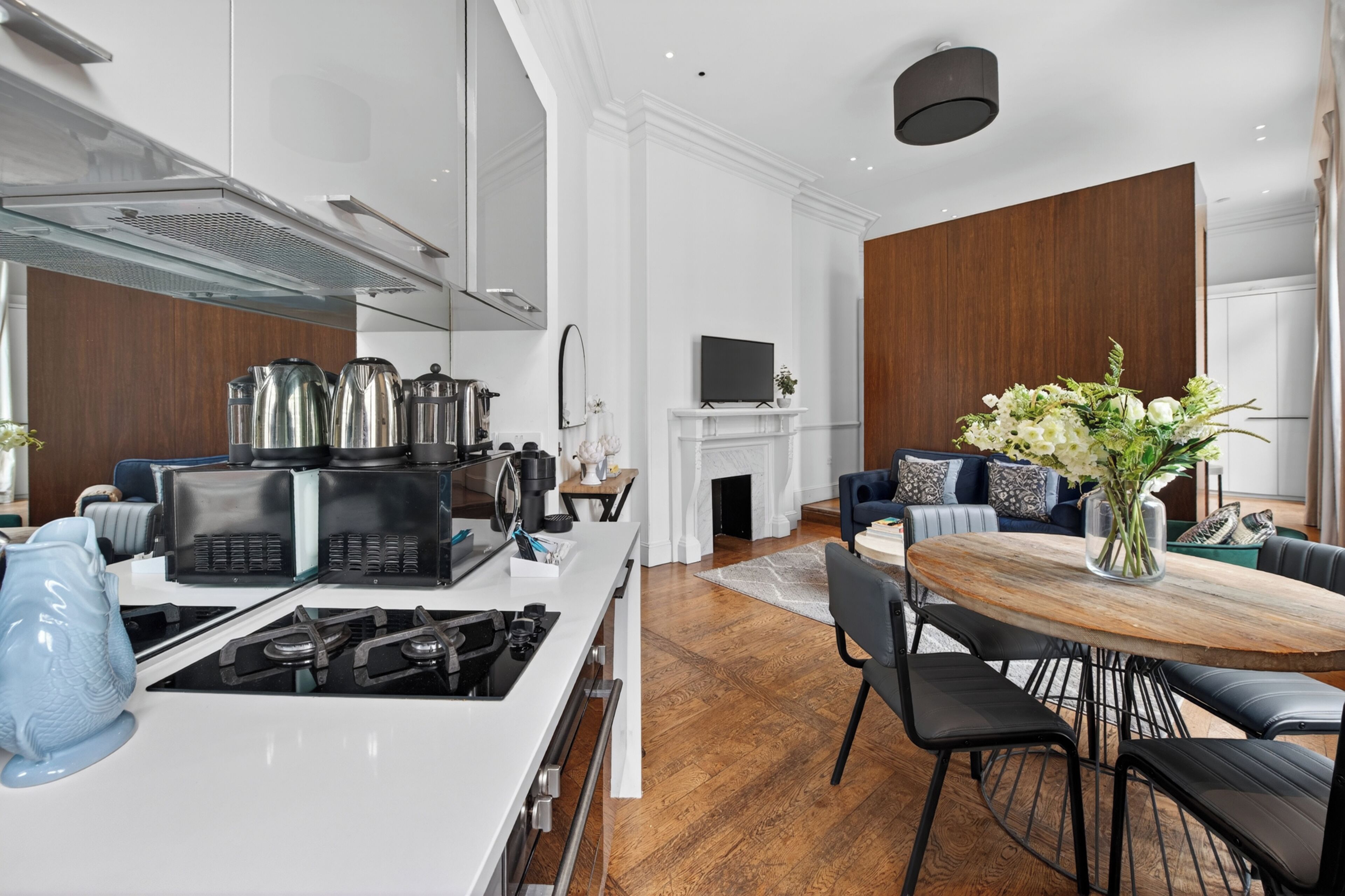 Uma cozinha moderna com eletrodomésticos de aço inoxidável, uma sala de jantar com mesa de madeira e cadeiras pretas, e uma sala de estar com lareira e televisão.