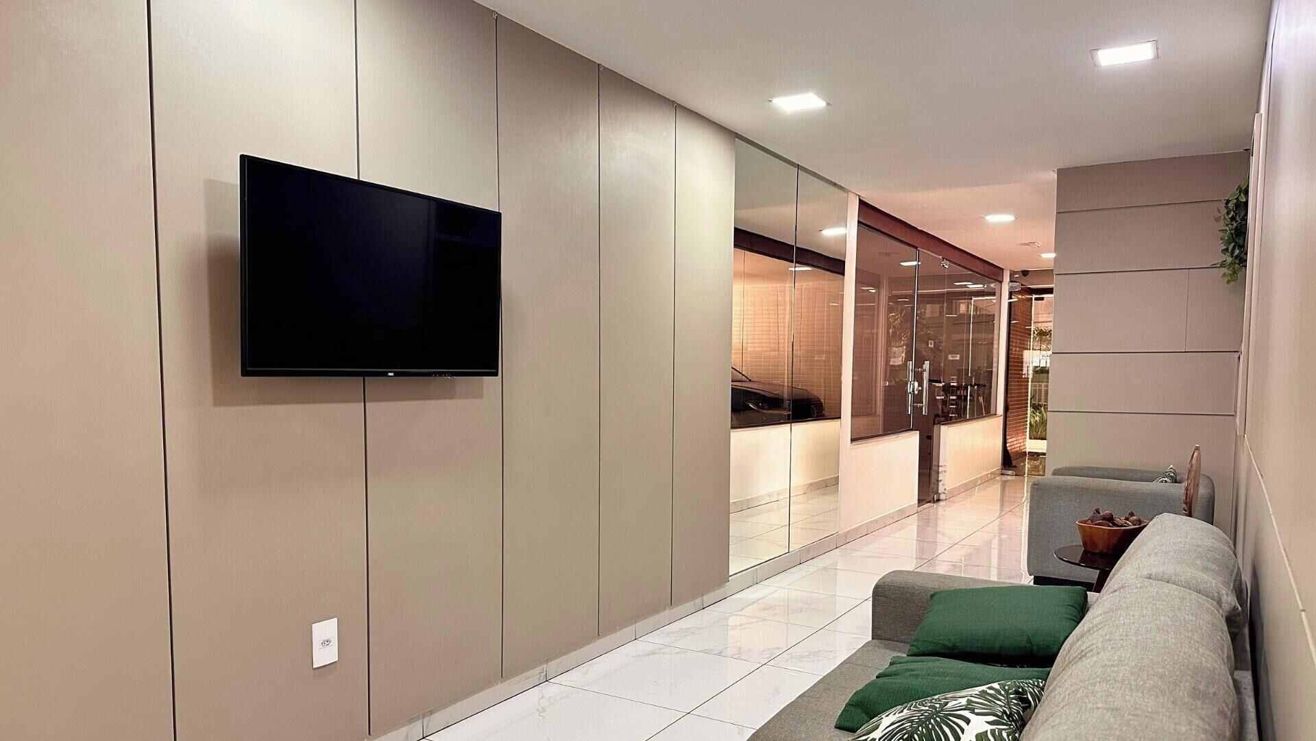 Um interior moderno com uma TV de tela plana na parede, um sofá com almofadas verdes e um painel de vidro separando a sala de estar da cozinha.