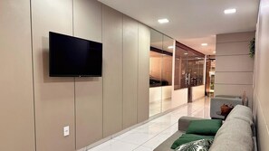 Um interior moderno com uma TV de tela plana na parede, um sofá com almofadas verdes e um painel de vidro separando a sala de estar da cozinha.