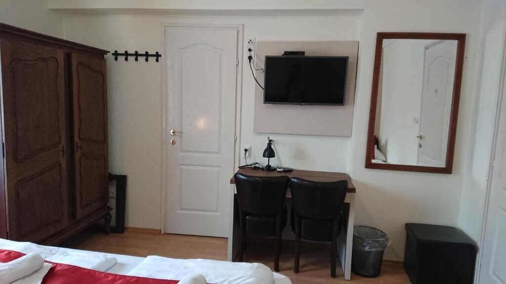 Quarto com uma cama, uma escrivaninha com duas cadeiras, televisão, guarda-roupa e espelho.