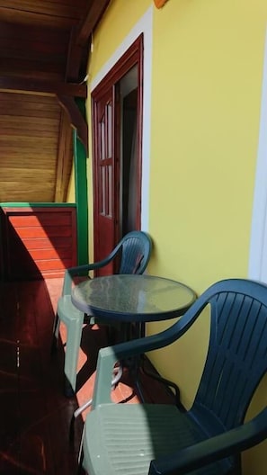 Uma área de estar coberta ao ar livre, com mesa redonda e duas cadeiras, teto de madeira e uma porta com moldura vermelha.