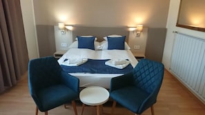 Quarto de hotel com uma cama, duas poltronas azuis, uma mesa redonda pequena e luminárias fixadas na parede.