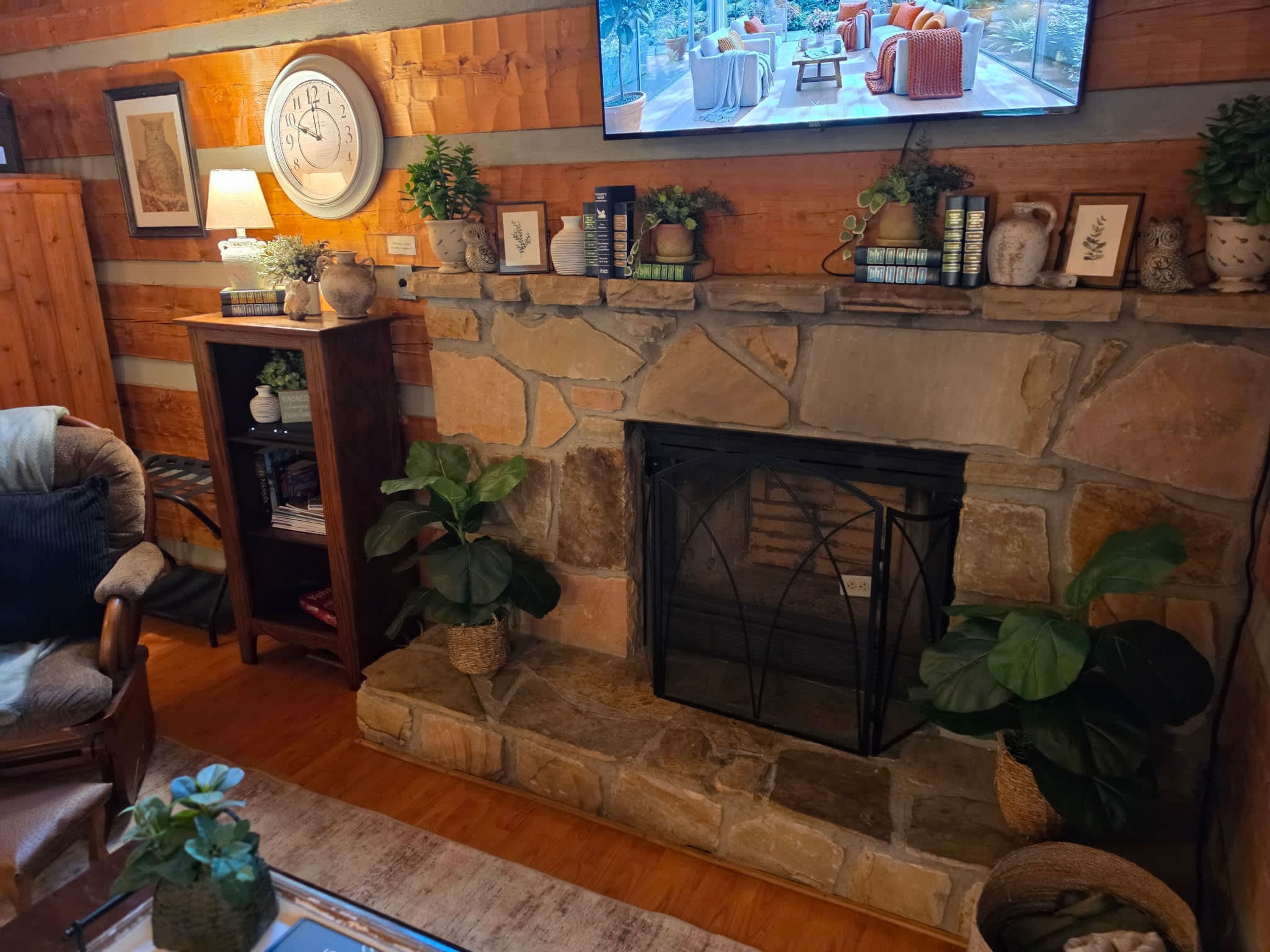 Uma sala de estar acolhedora com lareira de pedra, televisão montada acima e diversos objetos decorativos, incluindo livros e plantas.