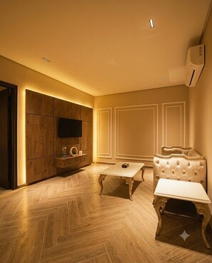 Quarto com piso de madeira, televisão fixada na parede, um sofá branco e uma mesa pequena.
