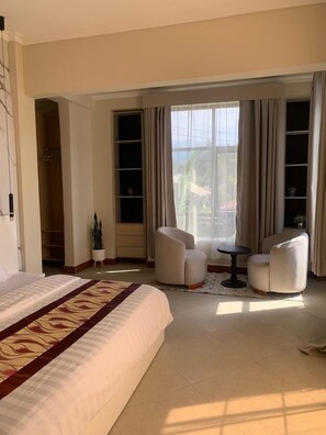 Quarto de hotel com uma cama grande, duas poltronas, uma mesinha, janela com cortinas e um guarda-roupa.