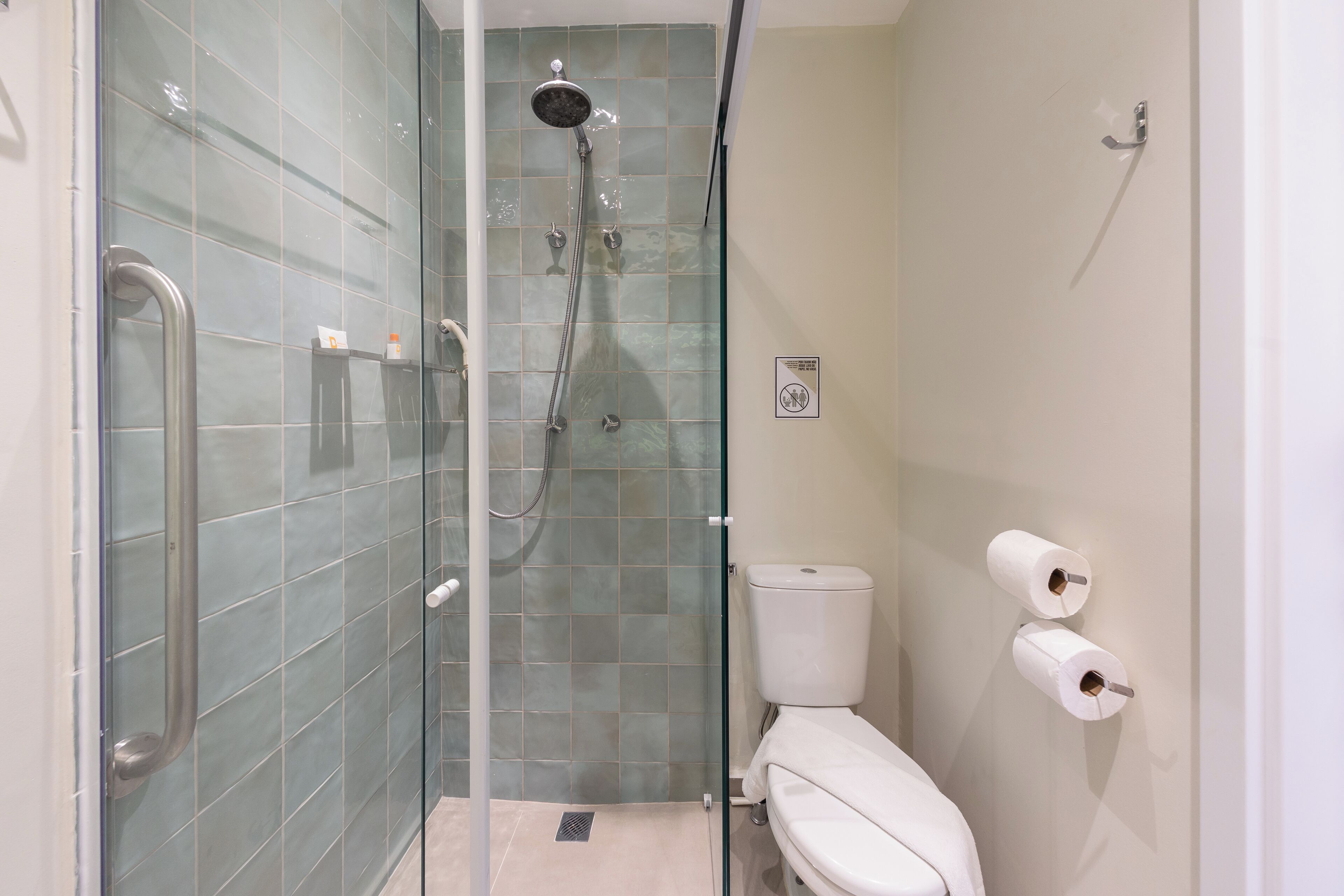 Uma casa de banho moderna com chuveiro de vidro, sanita branca e duas embalagens de papel higiénico na parede.