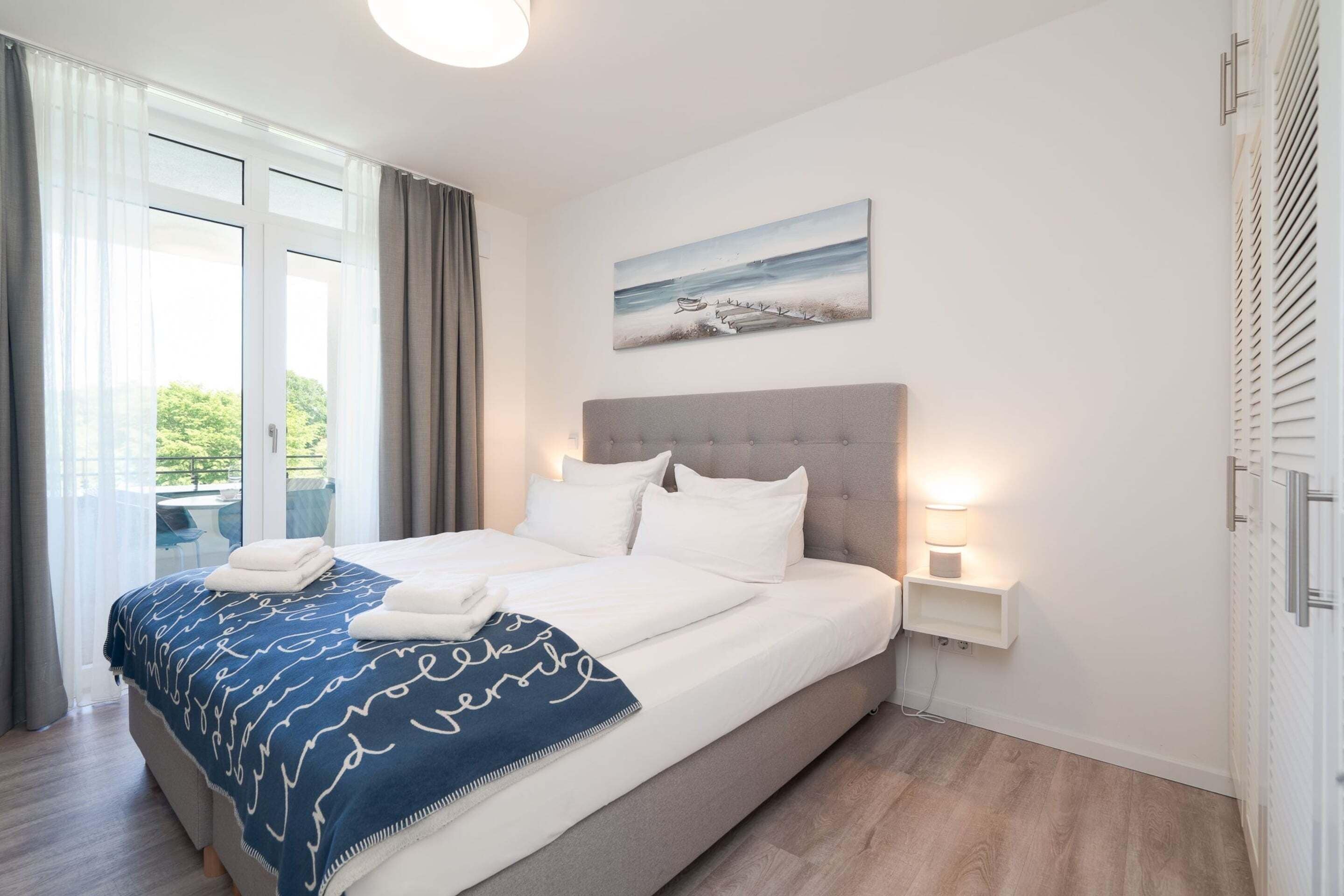 Quarto de hotel com uma cama, um quadro, uma janela com cortinas e vista para o verde lá fora.