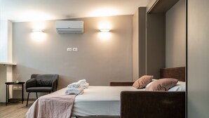 A modern bedroom with a bed, a chair, an air conditioning unit, and a bedside table.