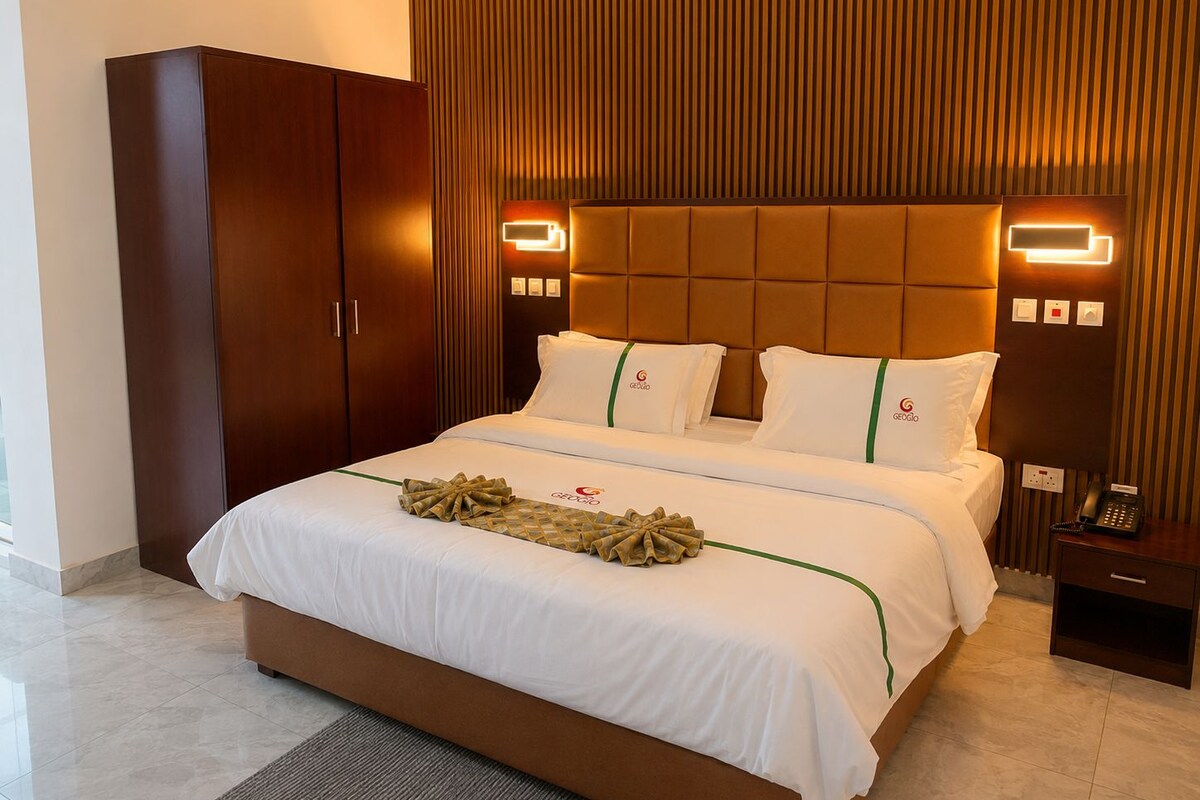 Quarto de hotel com uma cama grande, um guarda-roupa, mesa de cabeceira com telefone e luminária na parede.