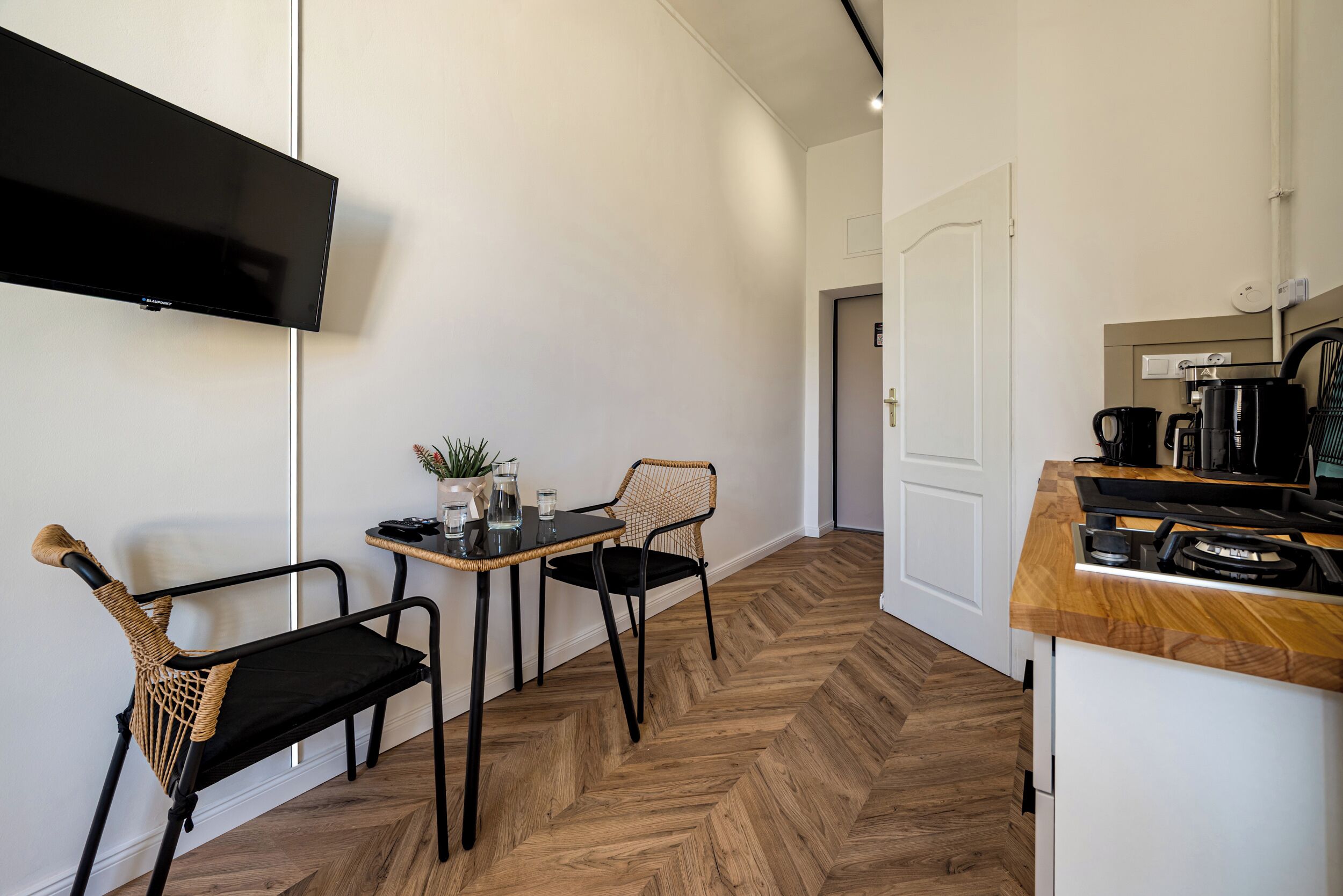 Uma cozinha compacta com piso de madeira em padrão herringbone, uma pequena área de jantar com duas cadeiras, uma mesa e uma TV de tela plana presa na parede.