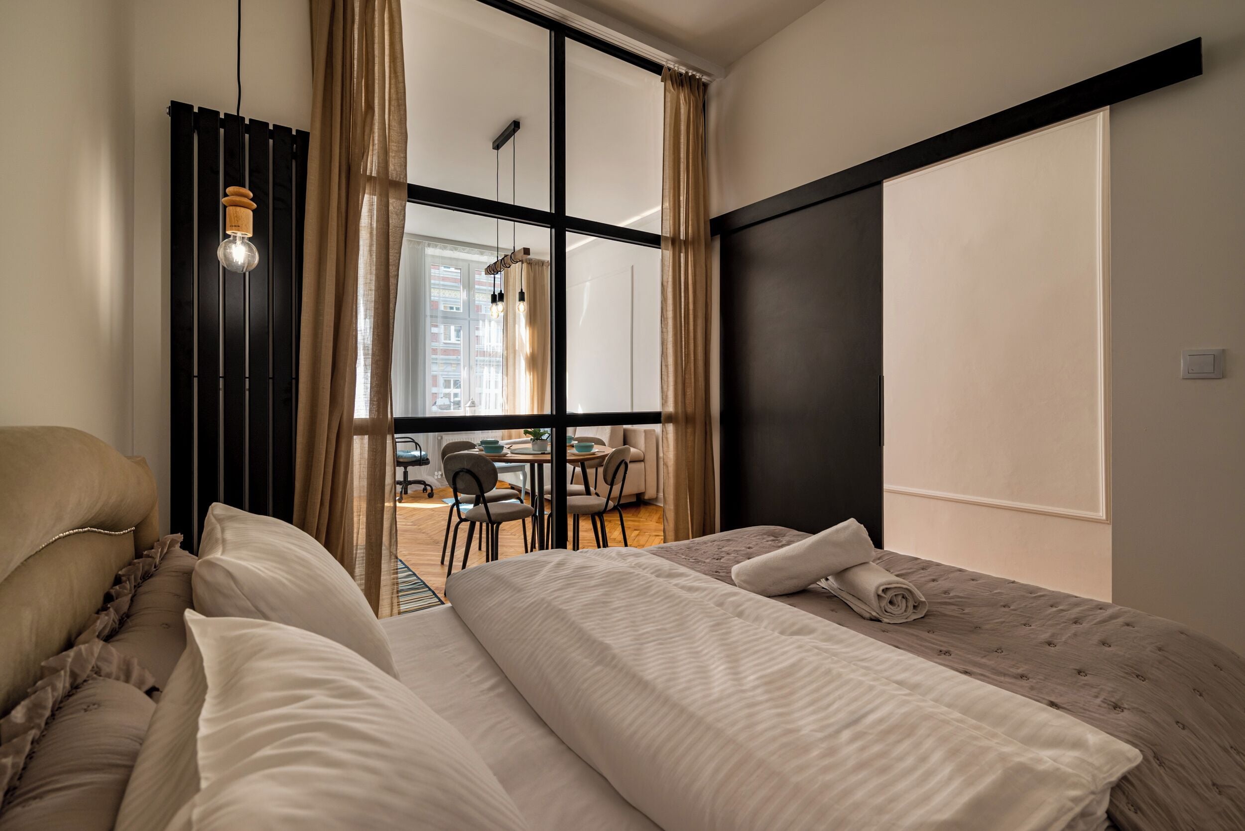 Quarto de hotel com cama, sofá, mesa de jantar com cadeiras e vista da cidade através de amplas janelas.